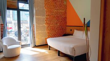 Quarto de hotel com cama, uma cadeira, janela com cortinas e parede de tijolos.