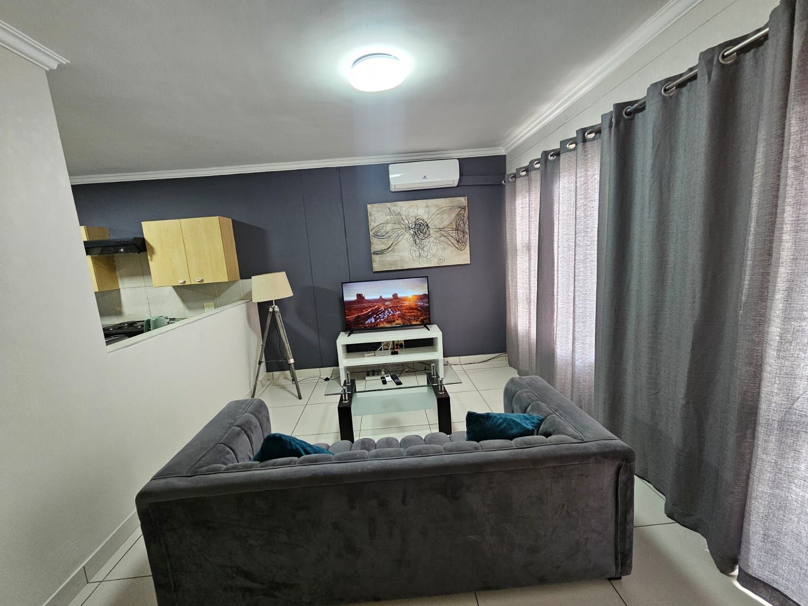 Uma sala de estar com um sofá cinza, uma televisão de tela plana, um abajur e um quadro na parede.