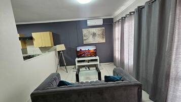 Uma sala de estar com um sofá cinza, uma televisão de tela plana, um abajur e um quadro na parede.