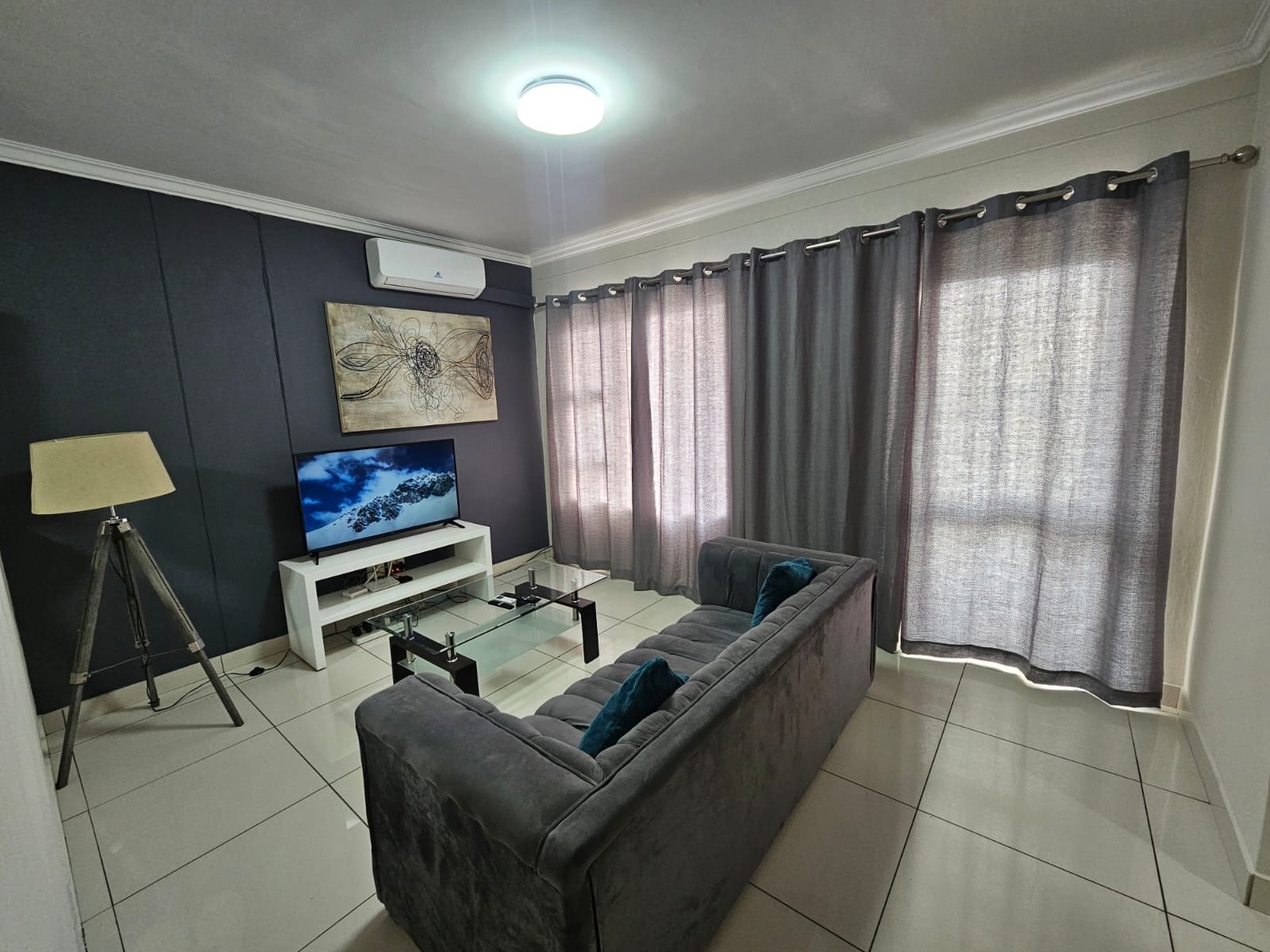 Uma sala de estar com sofá, televisão em um móvel, abajur e cortinas.