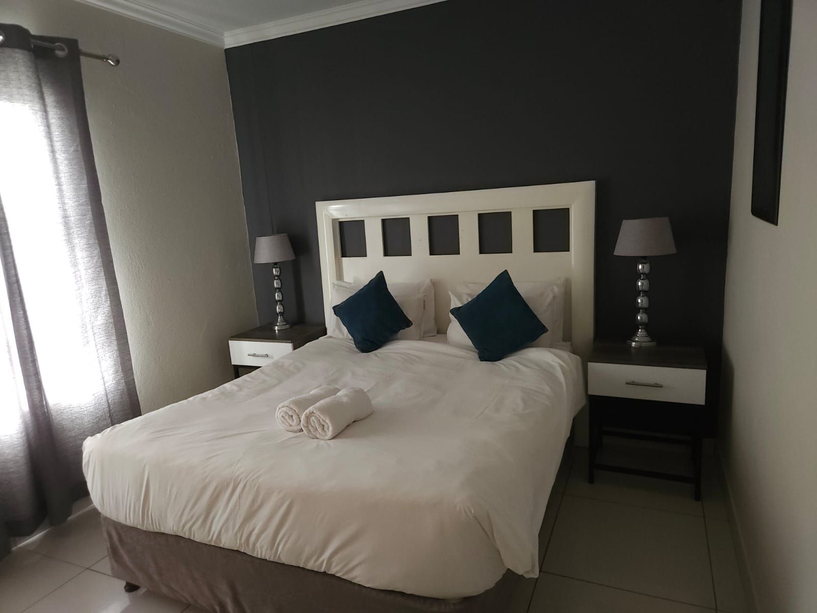 Quarto de hotel com cama, duas mesas de cabeceira com luminárias, janela com cortinas e uma parede escura.