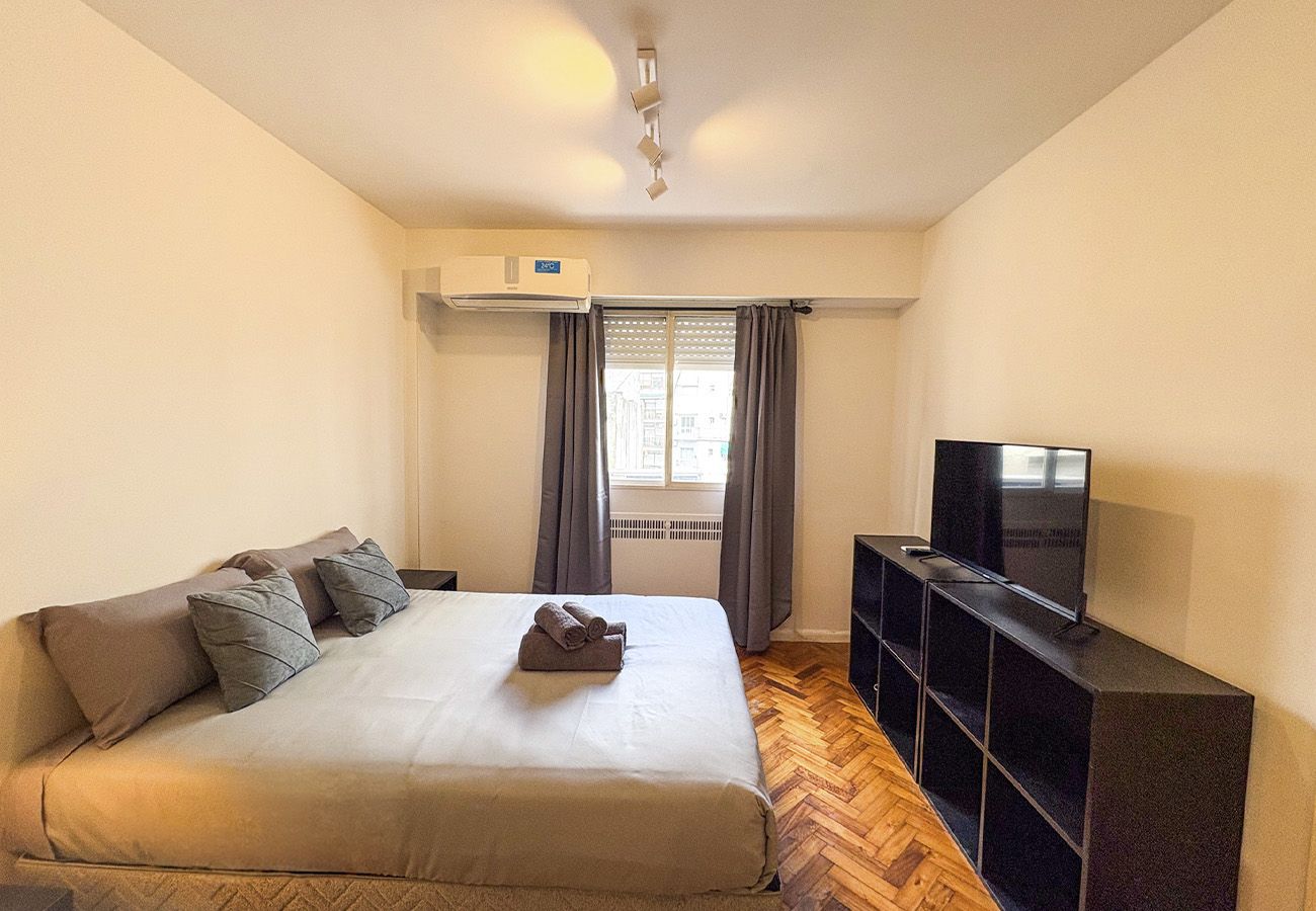 Quarto com cama, televisão sobre suporte, aparelho de ar condicionado e janela com cortinas.