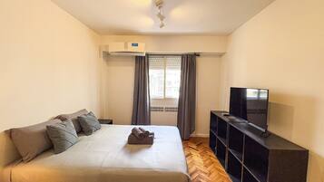Quarto com cama, televisão sobre suporte, aparelho de ar condicionado e janela com cortinas.