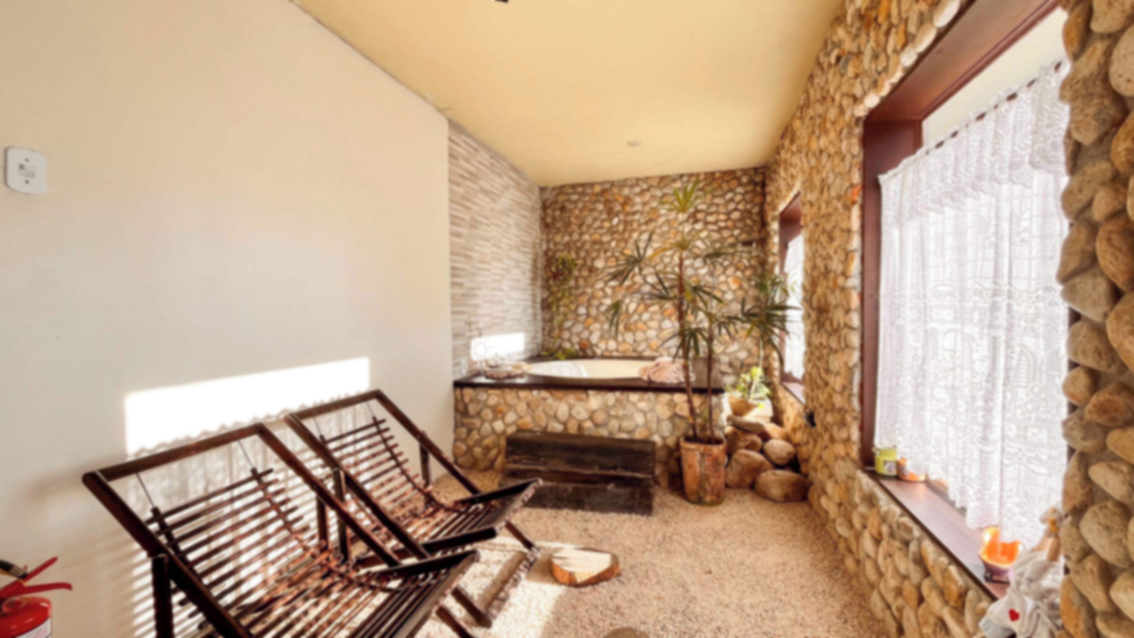 Quarto com parede de pedra, um banco de madeira, uma pequena mesa e janela com cortina.