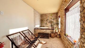 Quarto com parede de pedra, um banco de madeira, uma pequena mesa e janela com cortina.