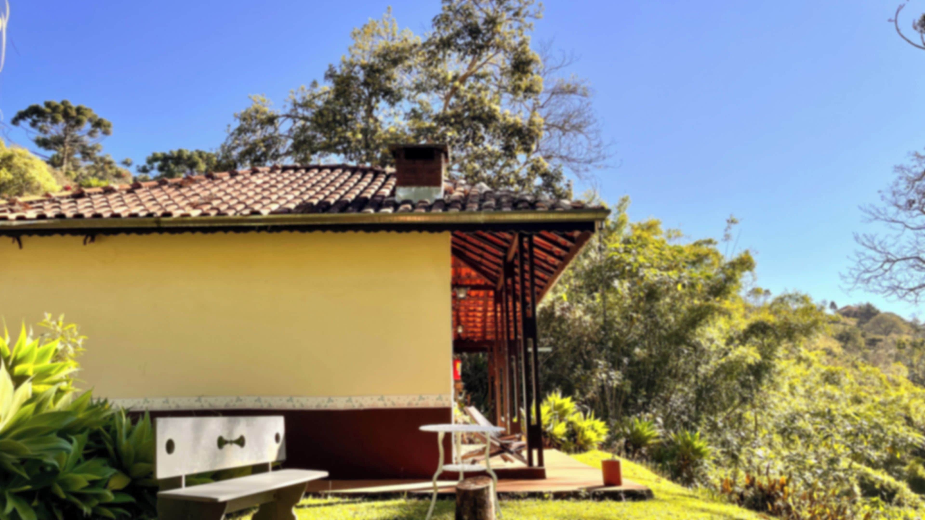 Um edifício amarelo com telhado de azulejos, área de estar coberta ao ar livre e vegetação exuberante.