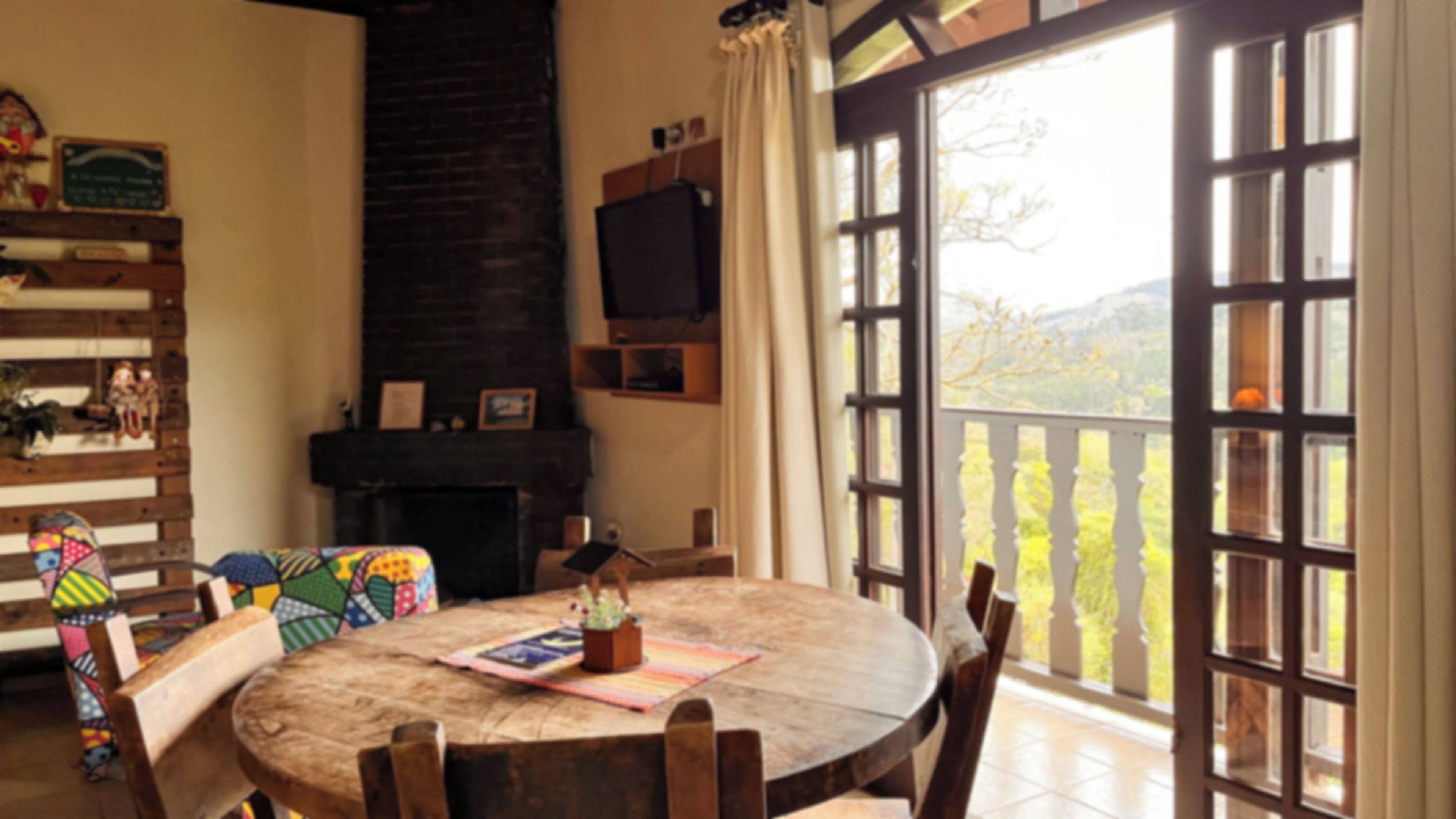 Mesa redonda de madeira com cadeiras, lareira e vista através de grandes janelas.