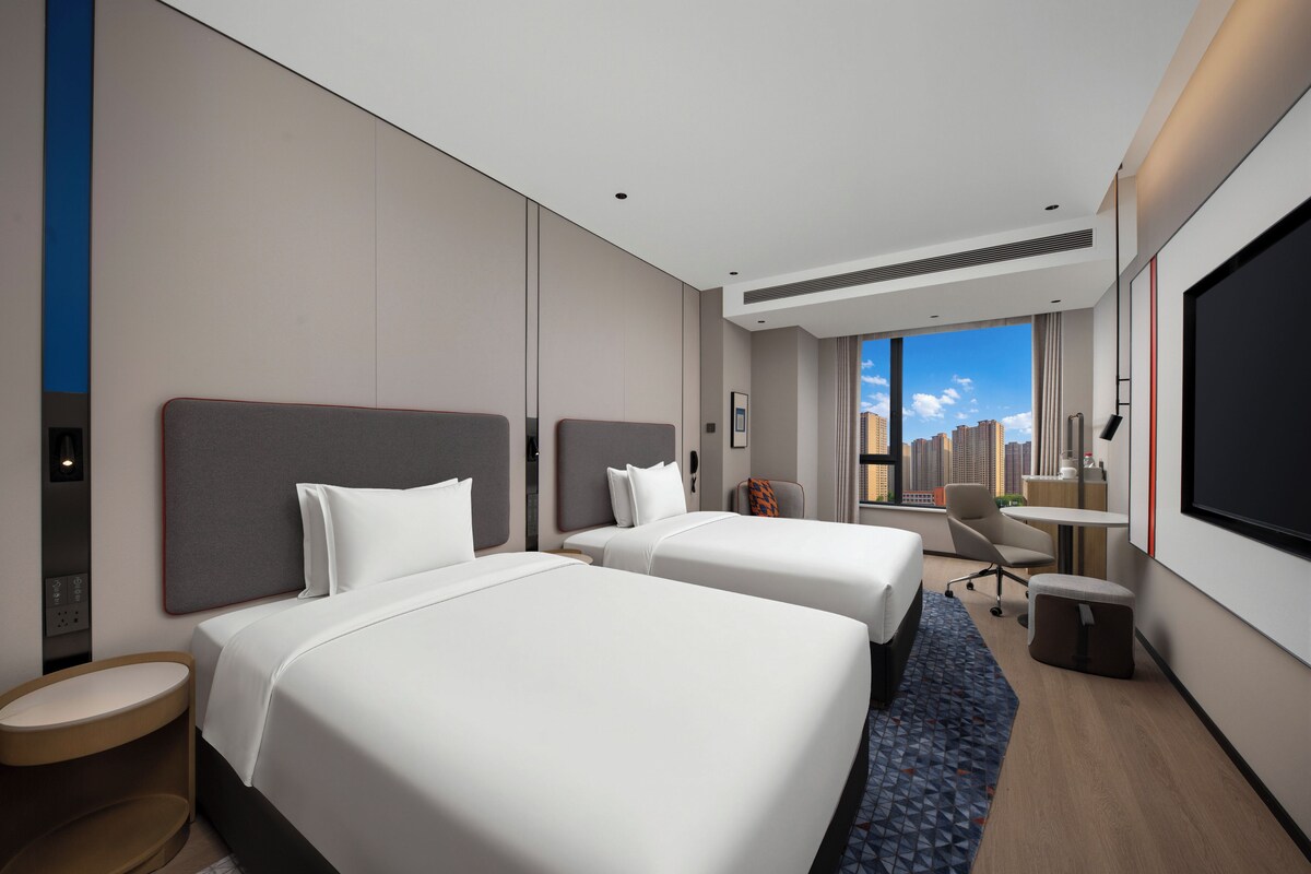 Quarto de hotel com duas camas, uma escrivaninha, uma cadeira, televisão e vista para a cidade.