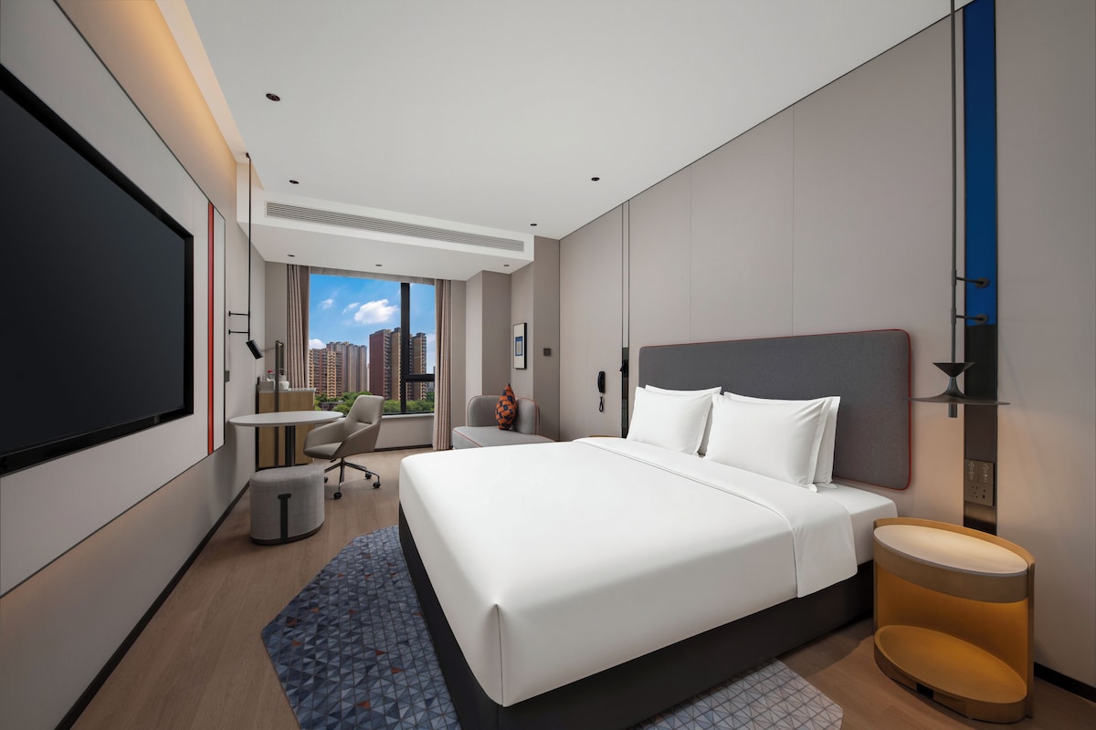 Quarto de hotel moderno com uma cama grande, uma escrivaninha e vista para a paisagem urbana.
