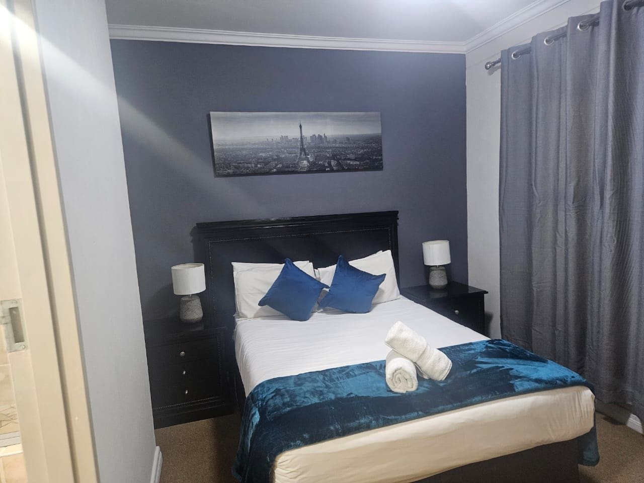 Quarto de hotel com cama, duas mesas de cabeceira, luminária, toalha e pintura da paisagem urbana na parede.