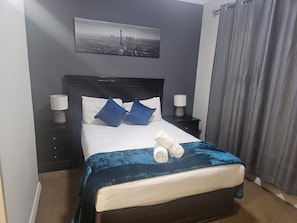 Cama bem arrumada com manta azul, duas almofadas e toalhas dobradas. Apresenta uma mesa de cabeceira com abajur e um quadro pendurado na parede.