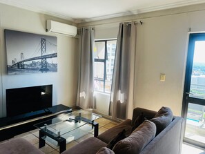 Uma sala de estar com televisão de tela plana, mesa de centro de vidro, sofá e uma janela grande com cortinas.