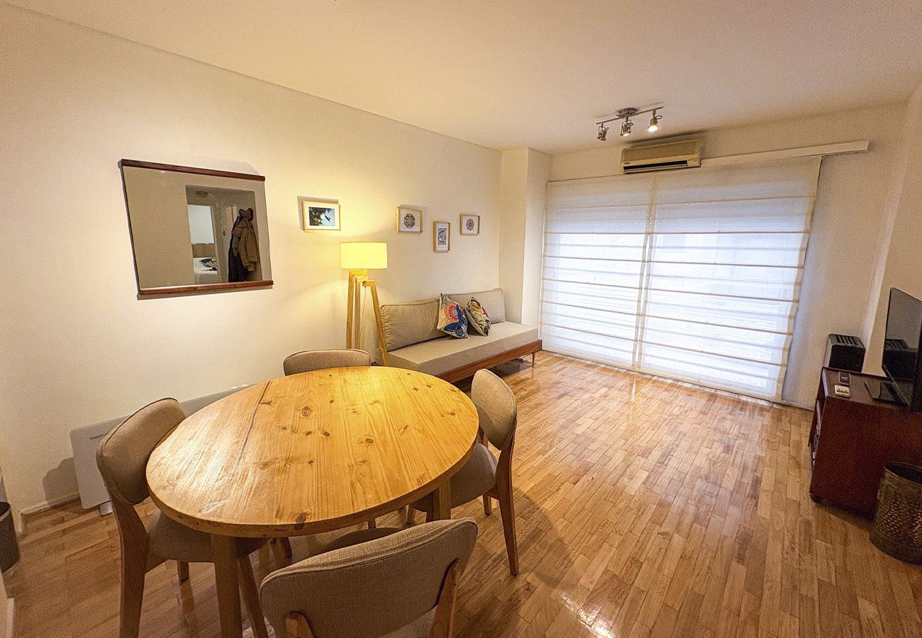 Quarto com mesa de madeira redonda, quatro cadeiras, sofá, luminária e televisão sobre um suporte.