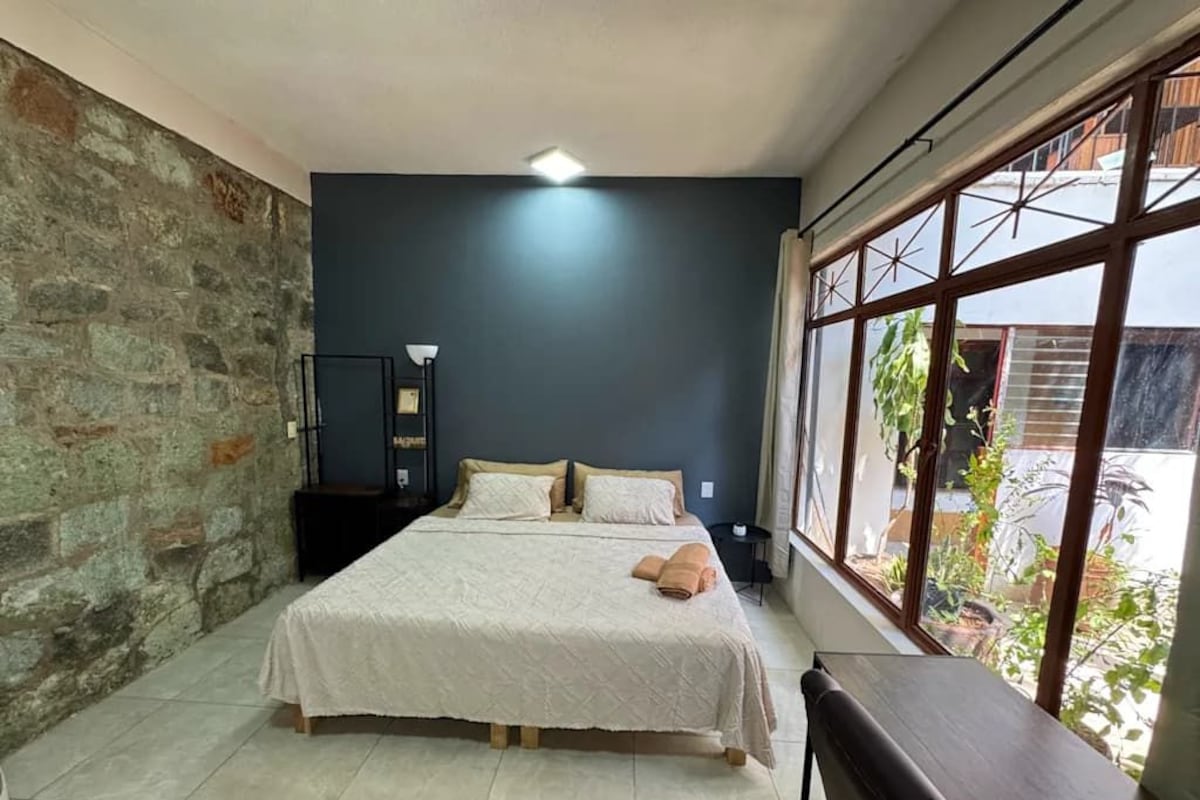 Quarto com cama grande, mesa de cabeceira, luminária e vista para o exterior pela janela.