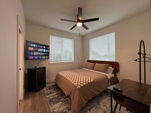 Quarto com cama, escrivaninha, televisão fixada na parede e ventilador de teto.