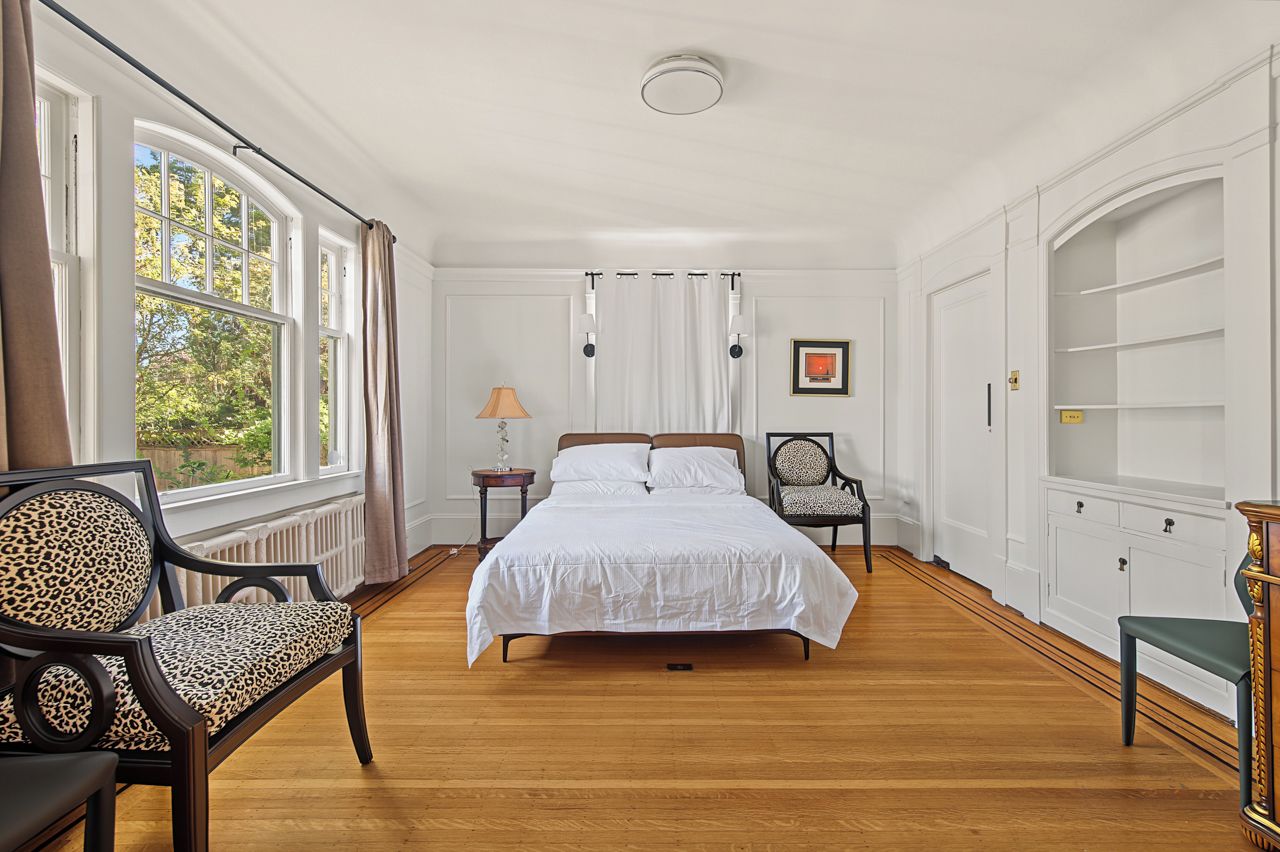 Quarto com cama grande, duas cadeiras, mesa de cabeceira e janela com cortinas.