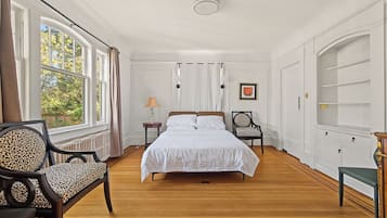 Quarto com cama grande, duas cadeiras, mesa de cabeceira e janela com cortinas.