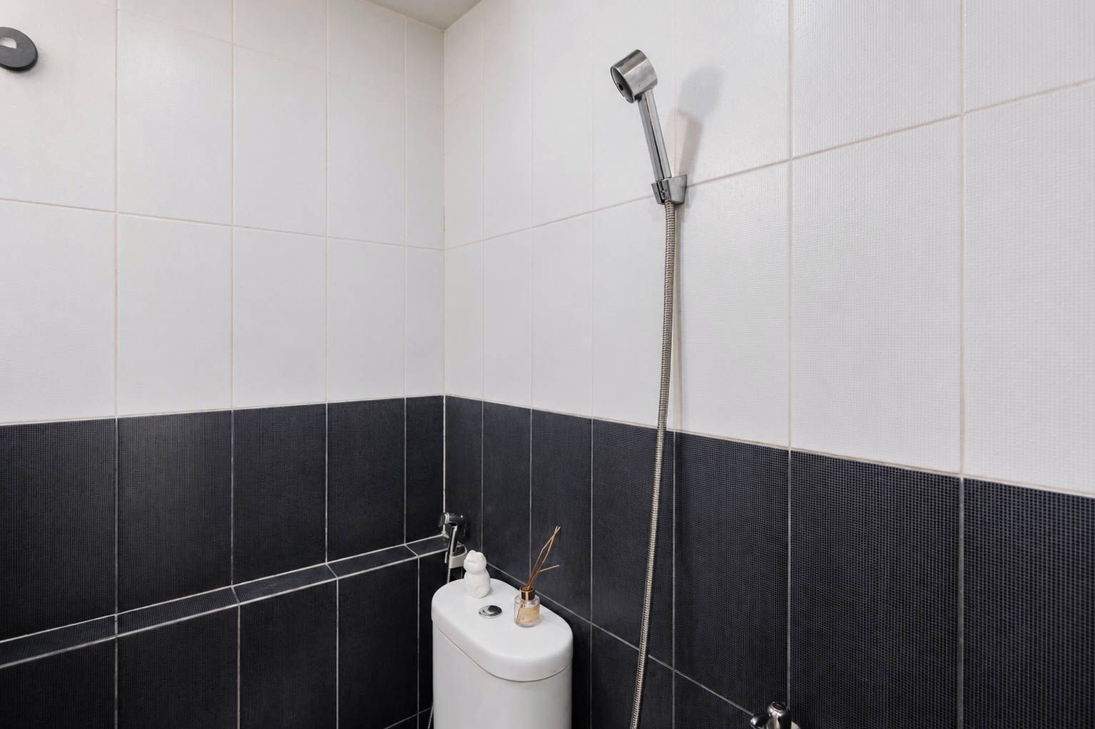 Casa de banho com azulejos brancos e negros, chuveiro com chuveiro manual e sanita com dispensador de sabão.
