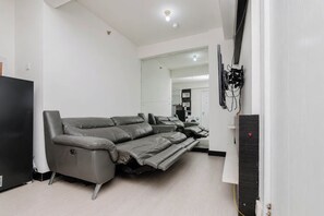 Uma sala de estar moderna com um sofá cinza, uma TV de tela plana na parede e uma máquina de lavar visível ao fundo.