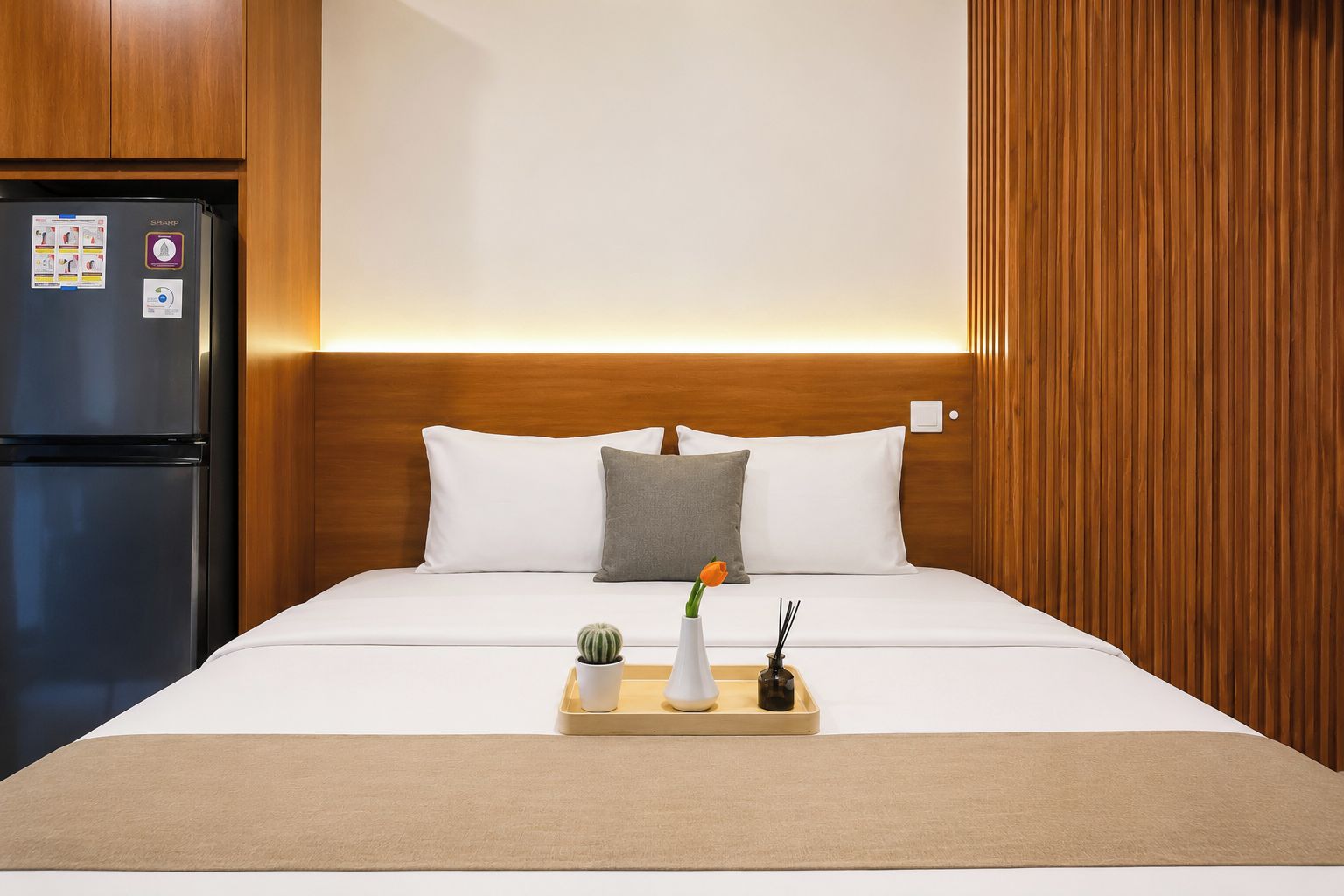 Quarto de hotel com cama, cabeceira de madeira, geladeira e bandeja com planta e vela.