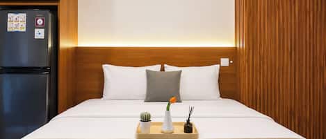 Quarto de hotel com cama, cabeceira de madeira, geladeira e bandeja com planta e vela.