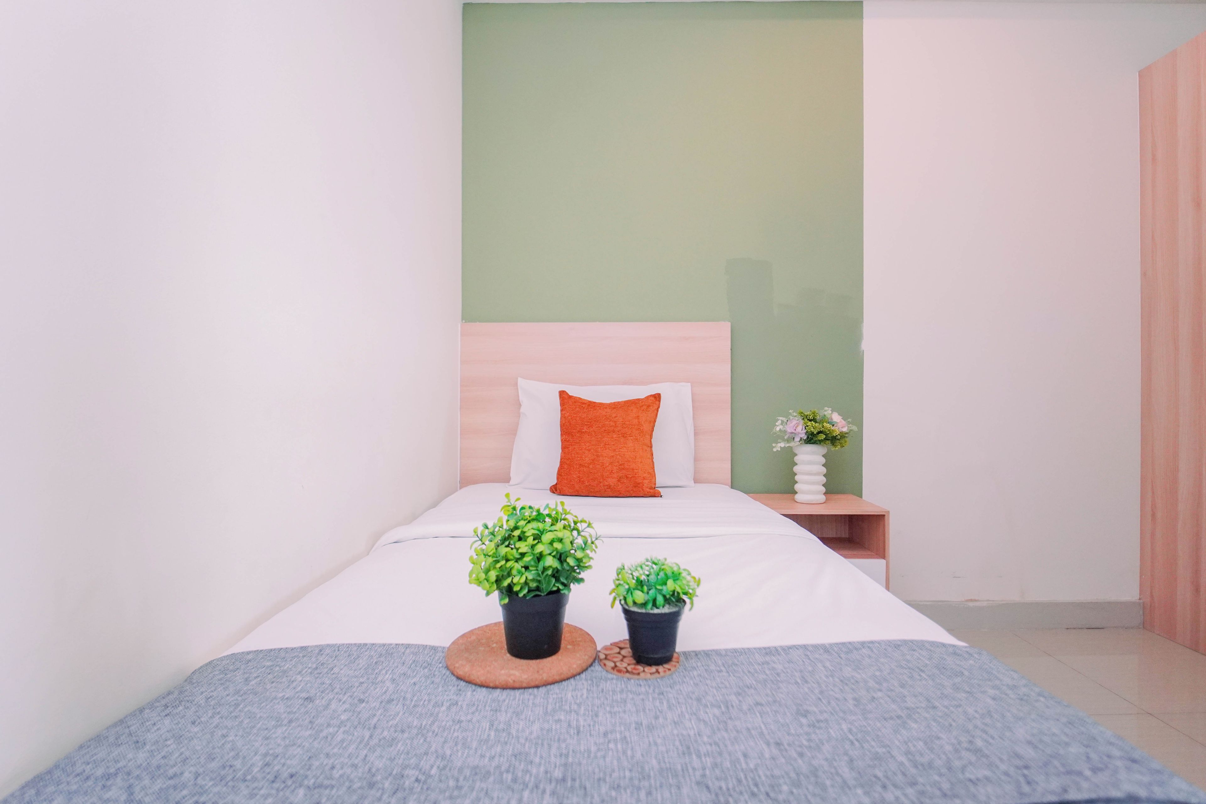 Cama bem arrumada com travesseiro branco e laranja, duas pequenas plantas em vasos e uma mesinha de cabeceira de madeira com um vaso de flores.