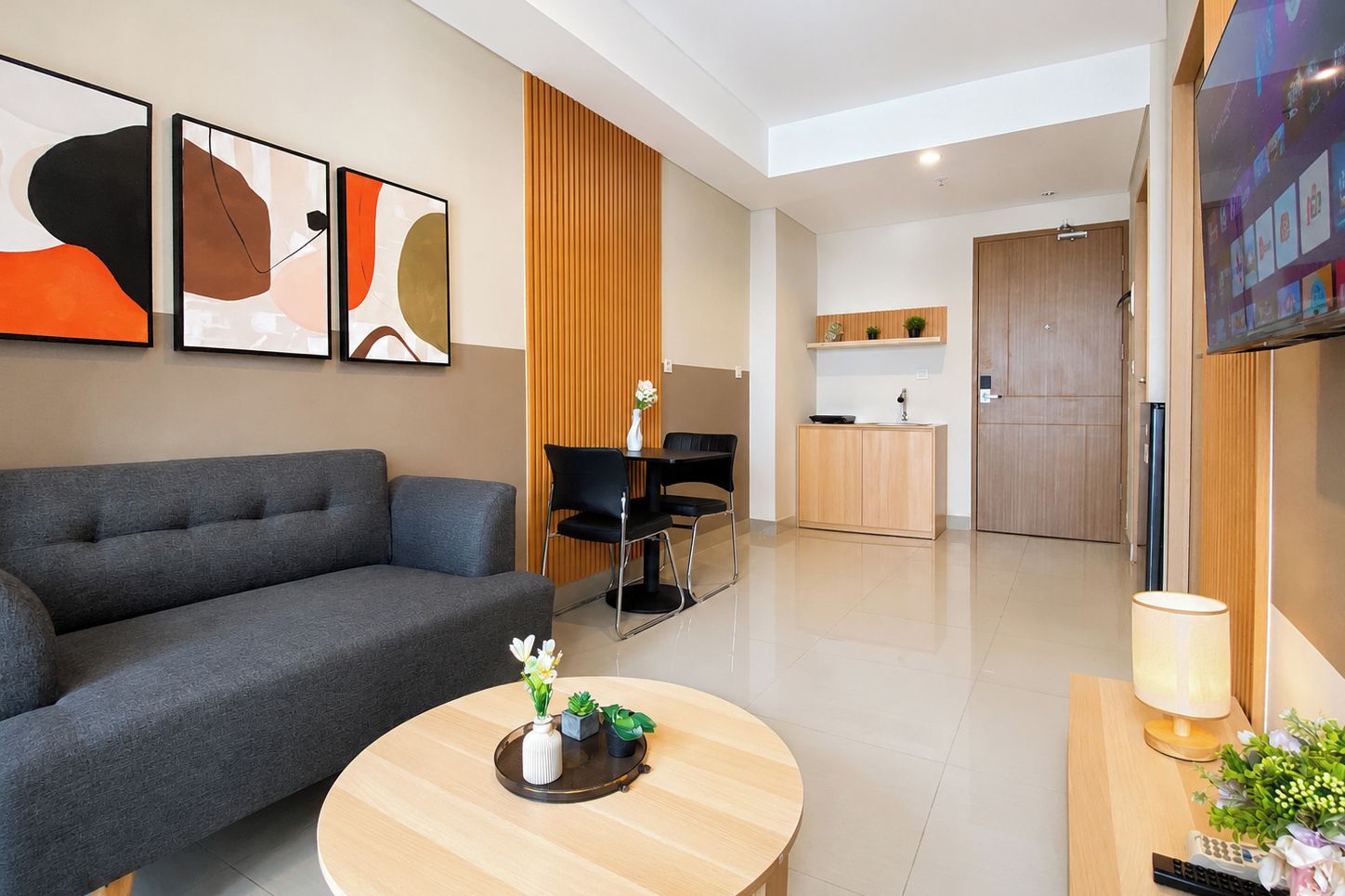 Uma sala de estar moderna com um sofá cinzento, uma mesa de centro redonda com objetos decorativos, uma televisão fixada na parede e uma área de cozinha ao fundo.