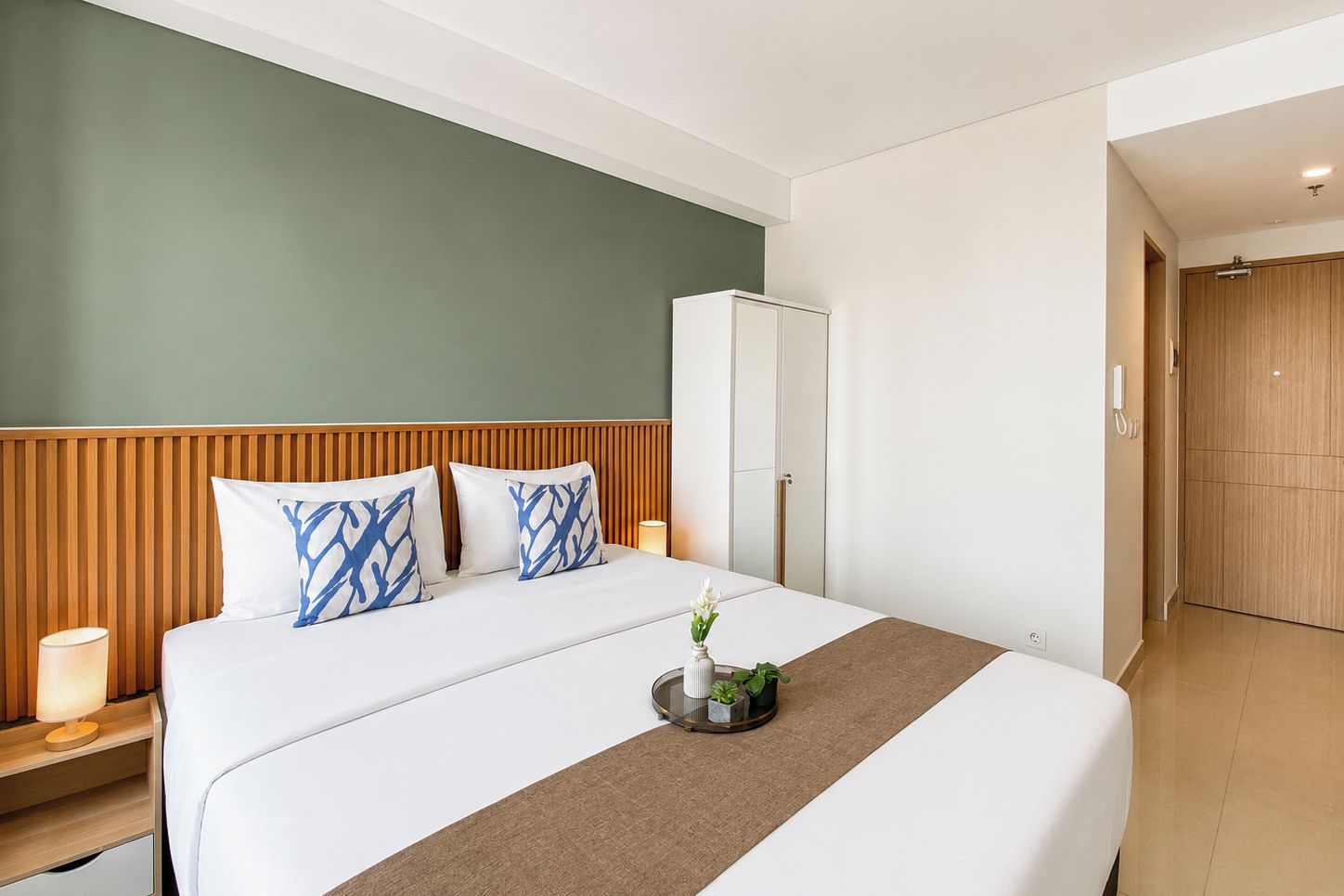 Quarto de hotel com cama, abajures, mesa de cabeceira, guarda-roupa e uma pequena planta em vaso sobre a cama.