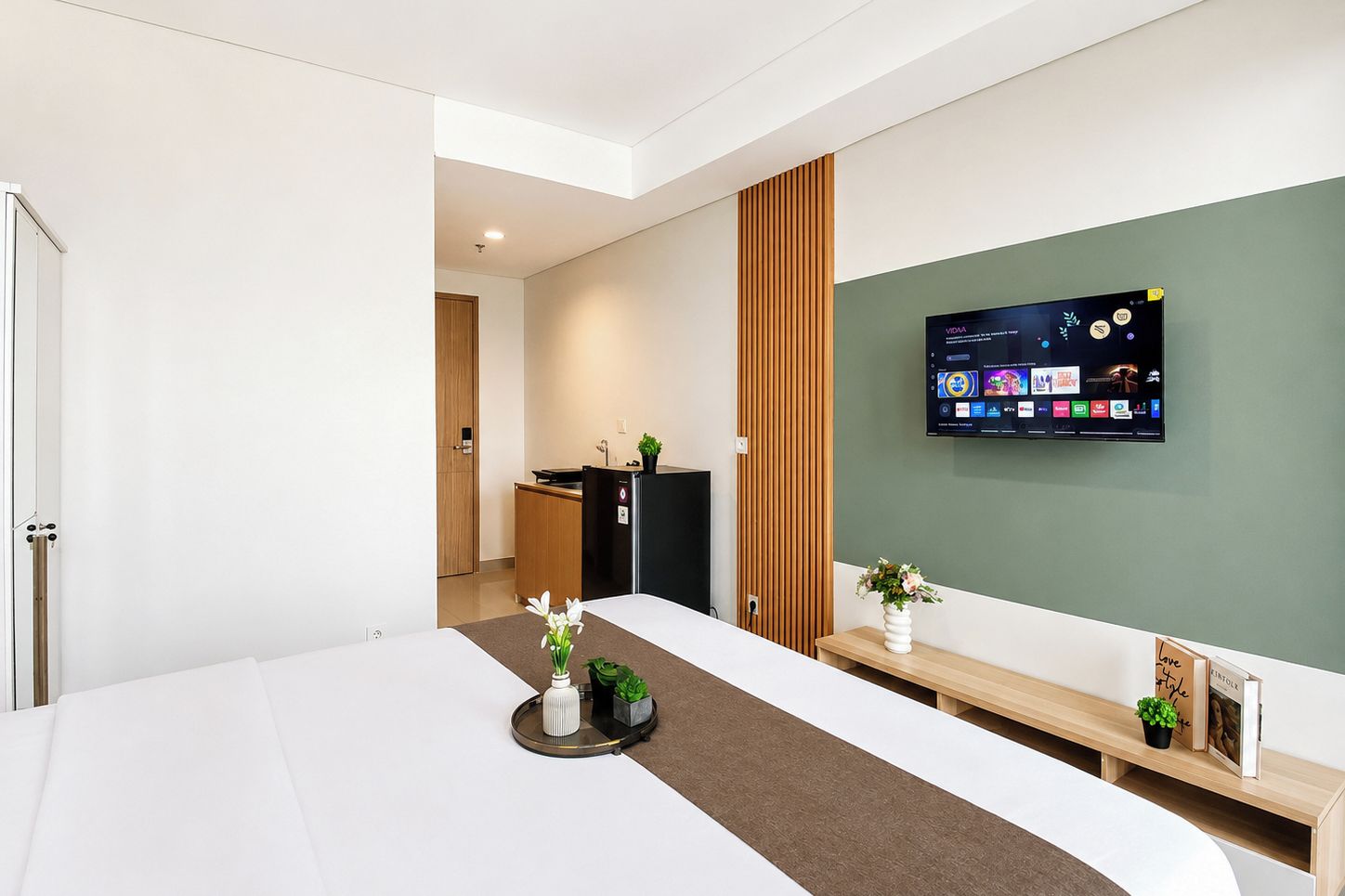 Quarto de hotel com uma cama grande, escrivaninha, televisão de tela plana na parede e uma pequena cozinha no fundo.