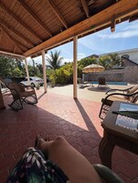 Área externa coberta com azulejos hexagonais, cadeiras de descanso, piscina e um guarda-sol de palha.