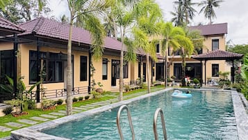 Piscina com borda de mármore, rodeada por palmeiras e um edifício com telhado em azulejos.