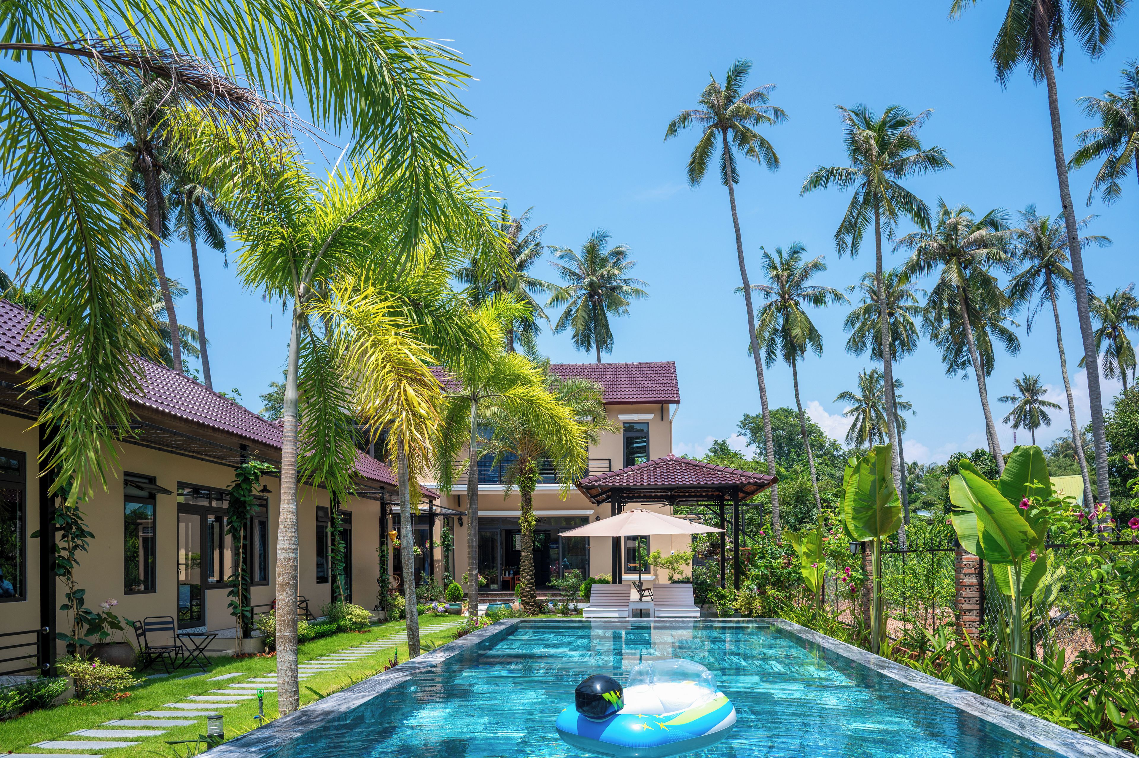 Área de piscina com piscina azul translúcida, rodeada por palmeiras e um edifício com telhado telhado.