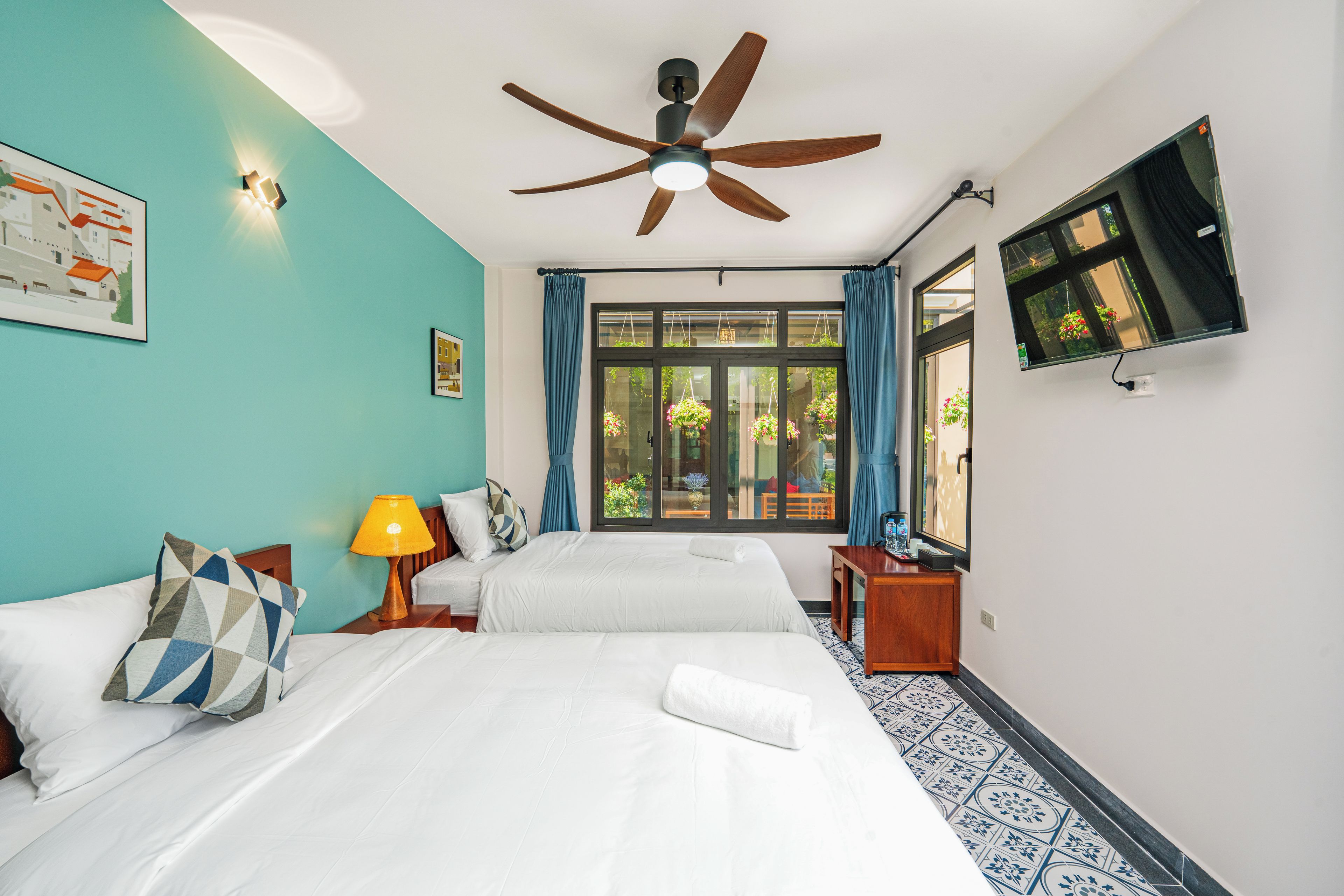 Quarto de hotel com duas camas, ventilador de teto, televisão fixada na parede e janela com cortinas.