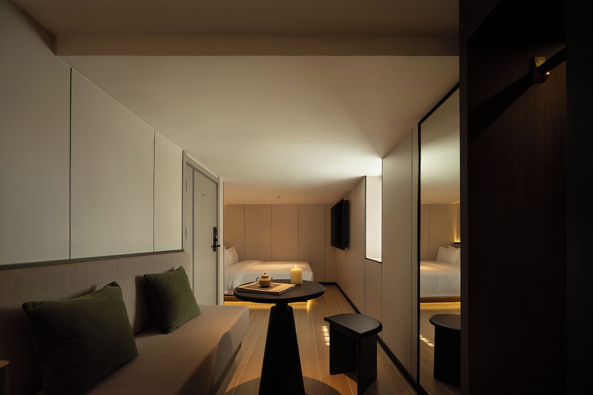 Quarto de hotel moderno com sofá, uma mesinha com vela e cama com TV na parede.