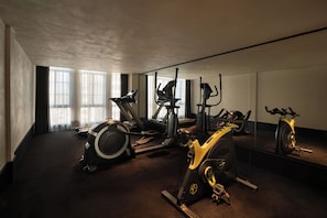 Academia com equipamentos de ginástica, amplas janelas e piso coberto por carpete.