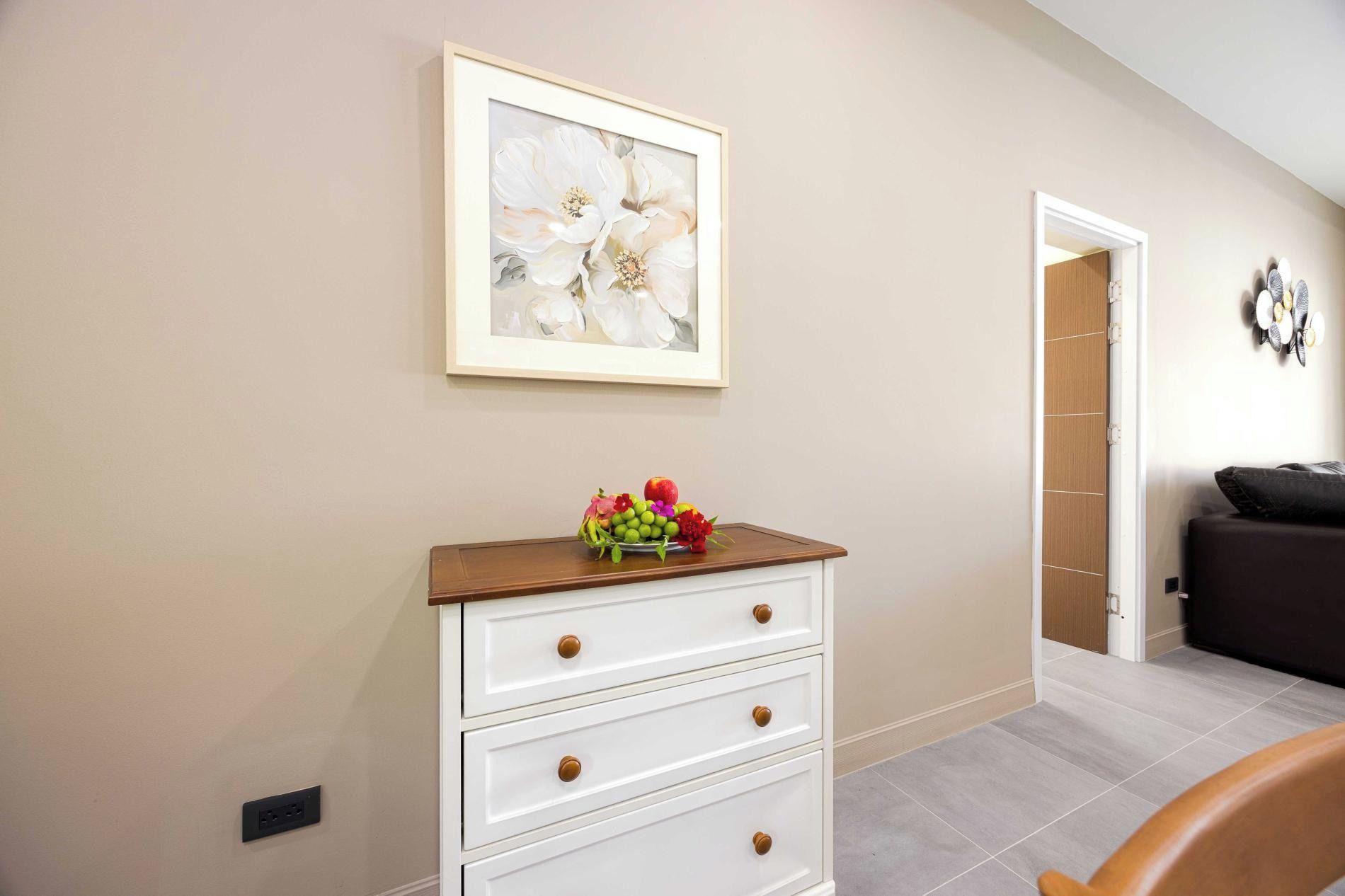 Quarto com penteadeira branca, uma pintura floral emoldurada na parede e uma porta que dá para outro cômodo.