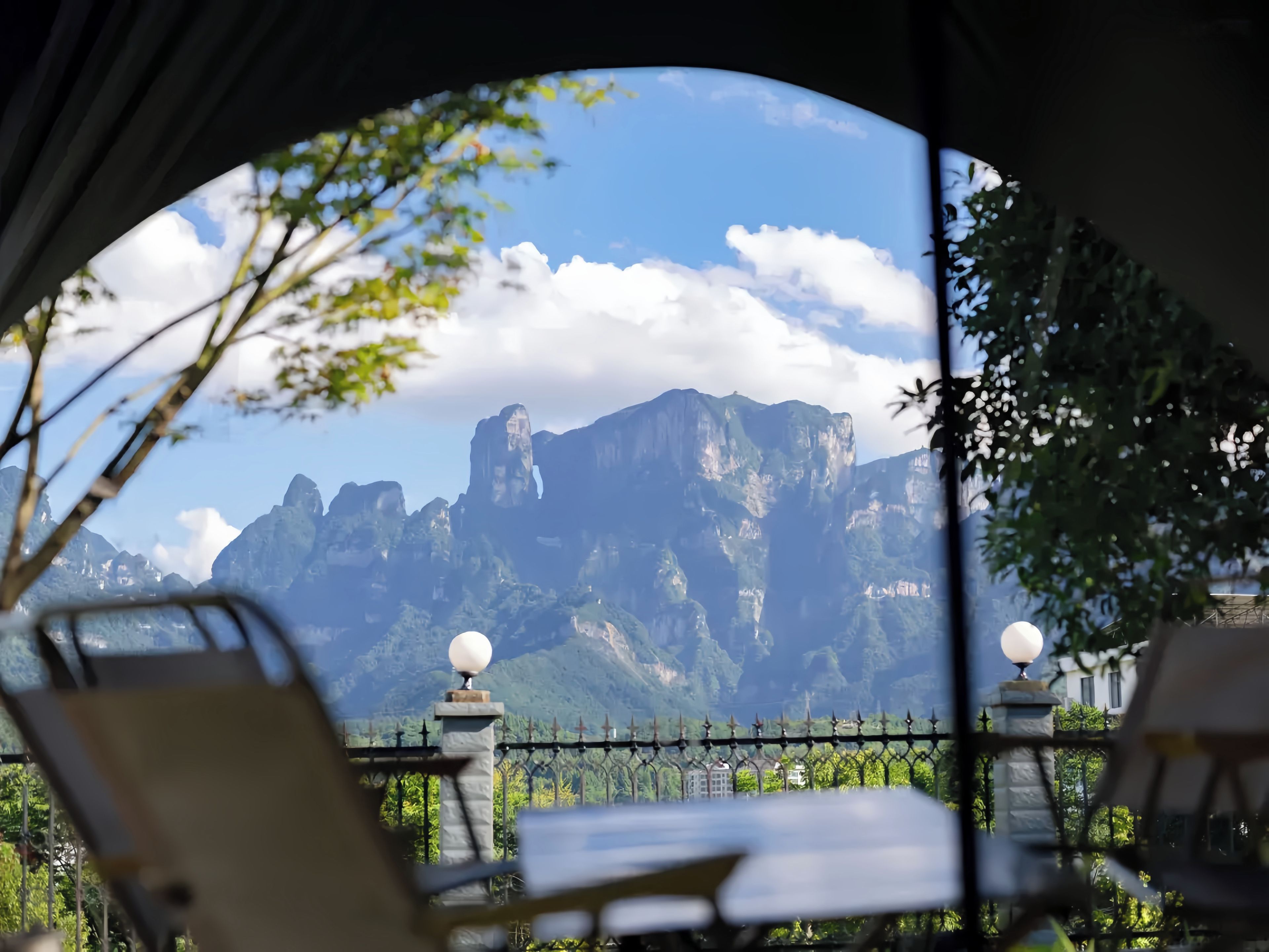 Vista através de uma janela curva de uma paisagem montanhosa, uma mesa e cadeiras com luminárias esféricas.
