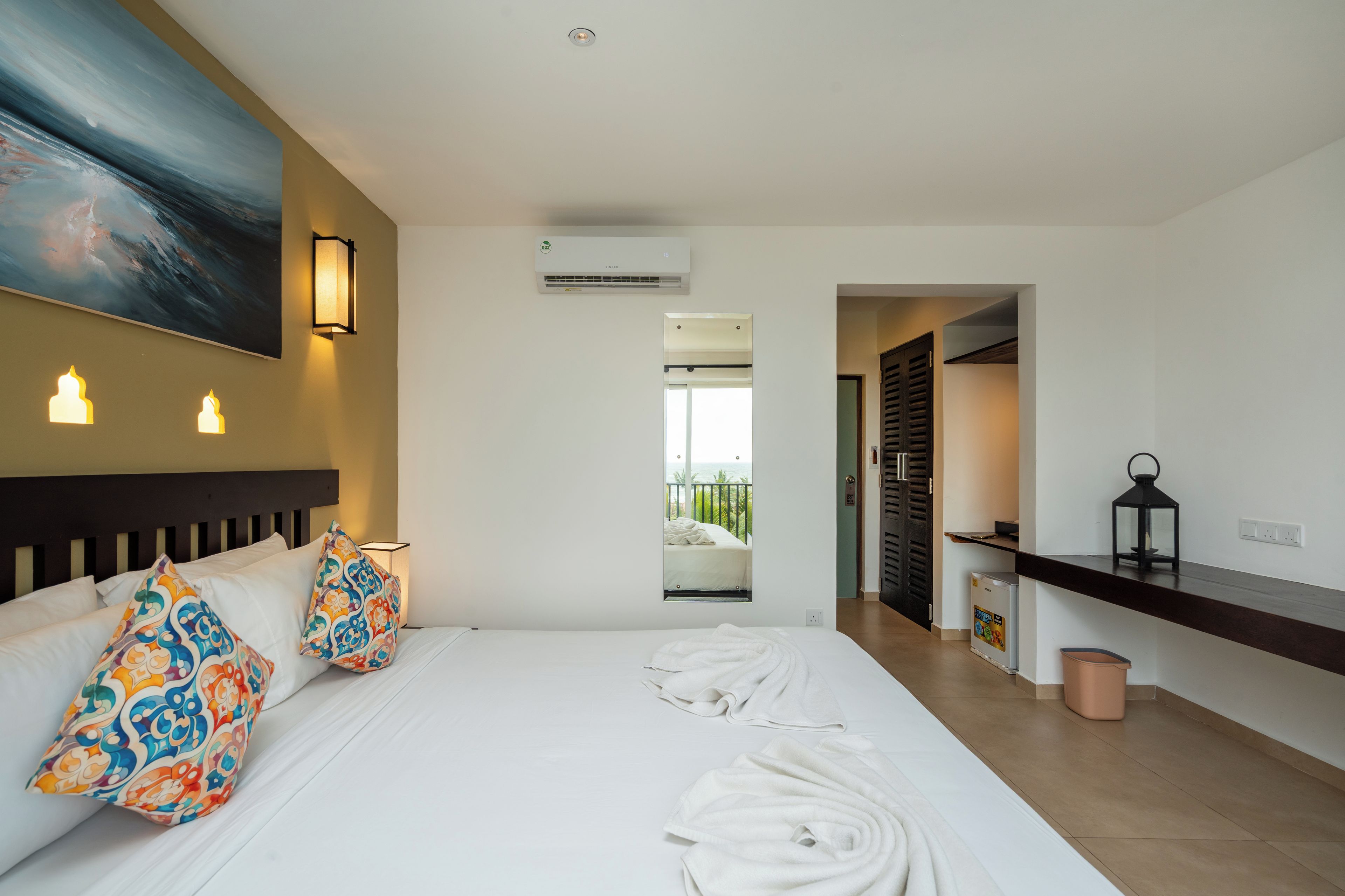 Quarto de hotel com cama, travesseiros, um quadro, mesa de cabeceira, espelho e vista para o mar.