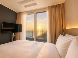 Quarto de hotel com cama, televisão, janela grande com cortinas e vista para o mar.