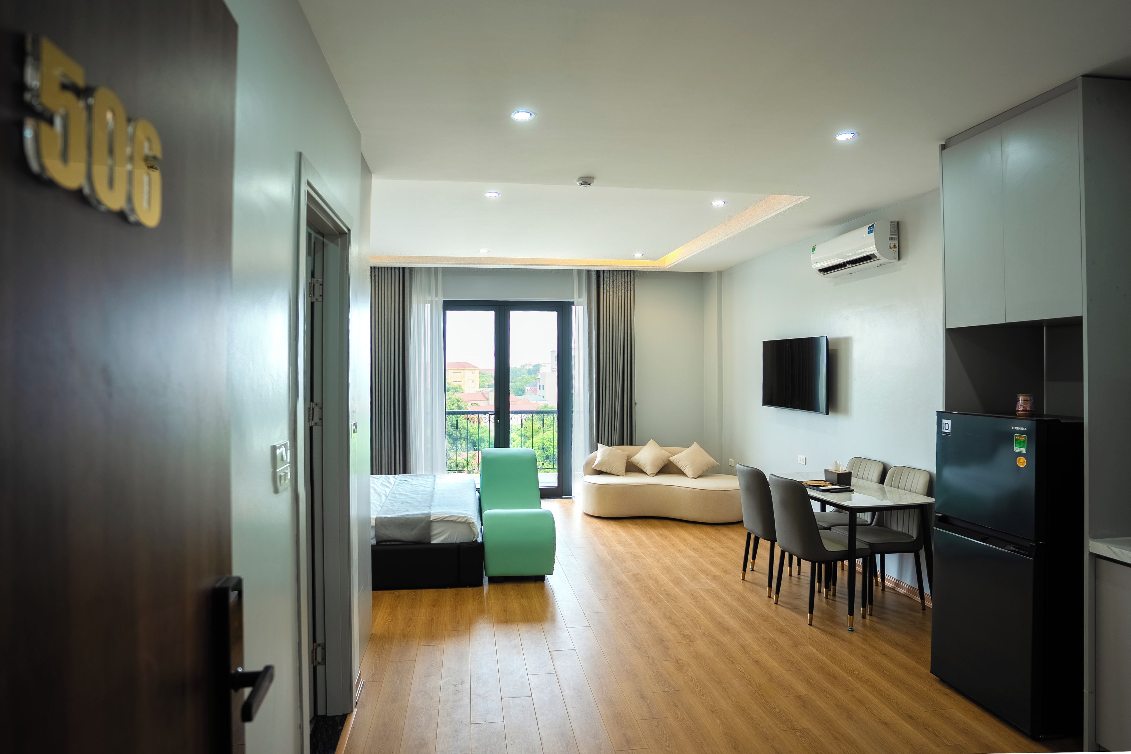 Quarto de hotel moderno com cama, sofá, mesa de jantar com cadeiras, televisão fixada na parede e aparelho de ar condicionado.