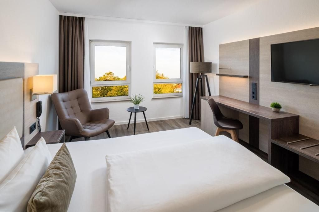 Quarto de hotel com cama, escrivaninha, cadeira, televisão e janelas com vista.