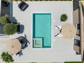 Vista aérea de uma piscina no terraço, com espreguiçadeiras e guarda-sóis ao redor, cercada por vegetação e um edifício.