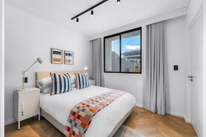 Quarto moderno com cama, mesas de cabeceira, janela com cortinas e uma obra de arte na parede.