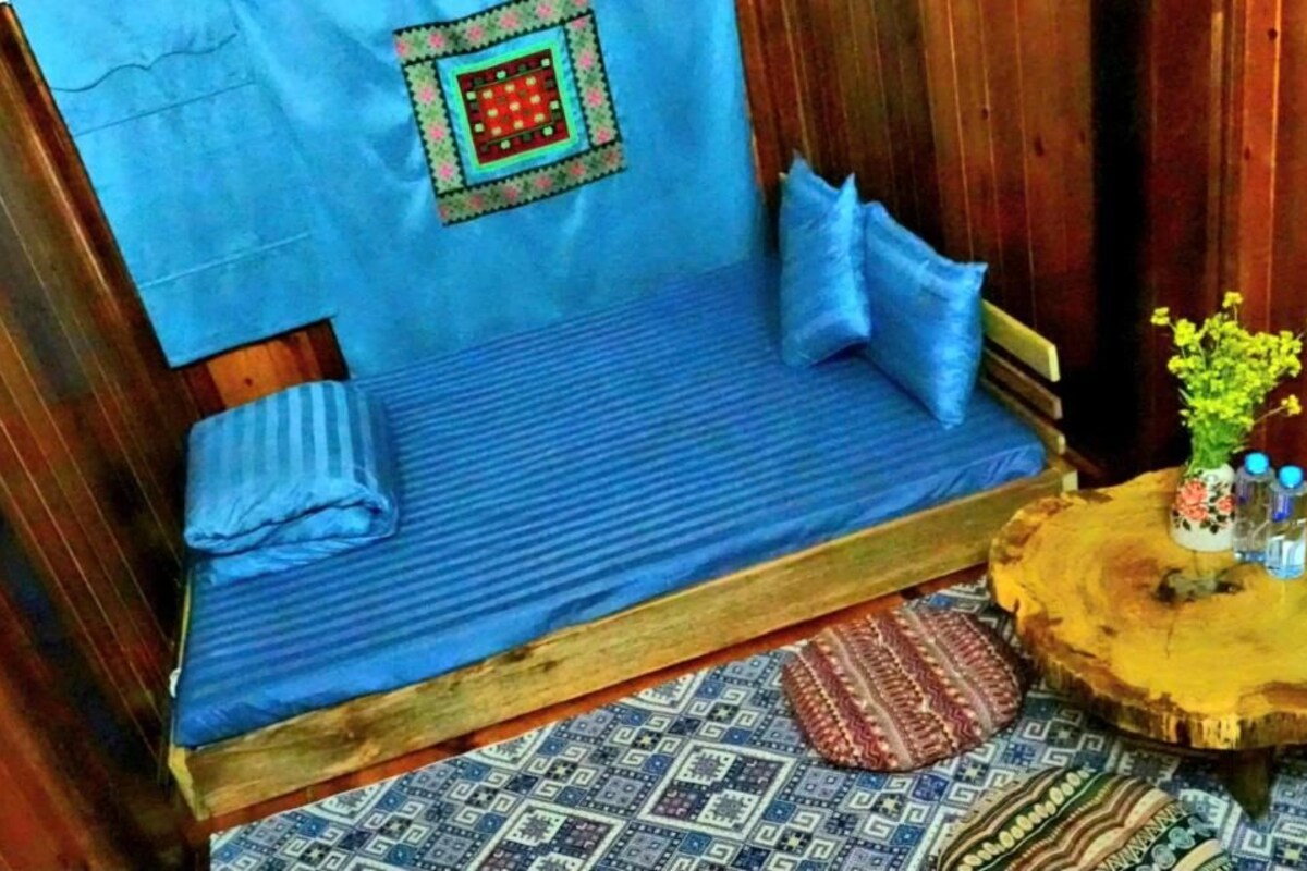 Um quarto aconchegante com um sofá azul, almofadas, uma pequena mesa de madeira com um vaso e tapetes estampados.
