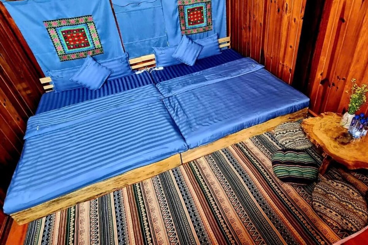 Quarto com cama, almofadas, mesa de cabeceira de madeira e tapete estampado.