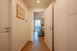 Corredor com portas brancas, uma partitura musical emoldurada na parede e vista para uma sala de estar com sofá e mesa.