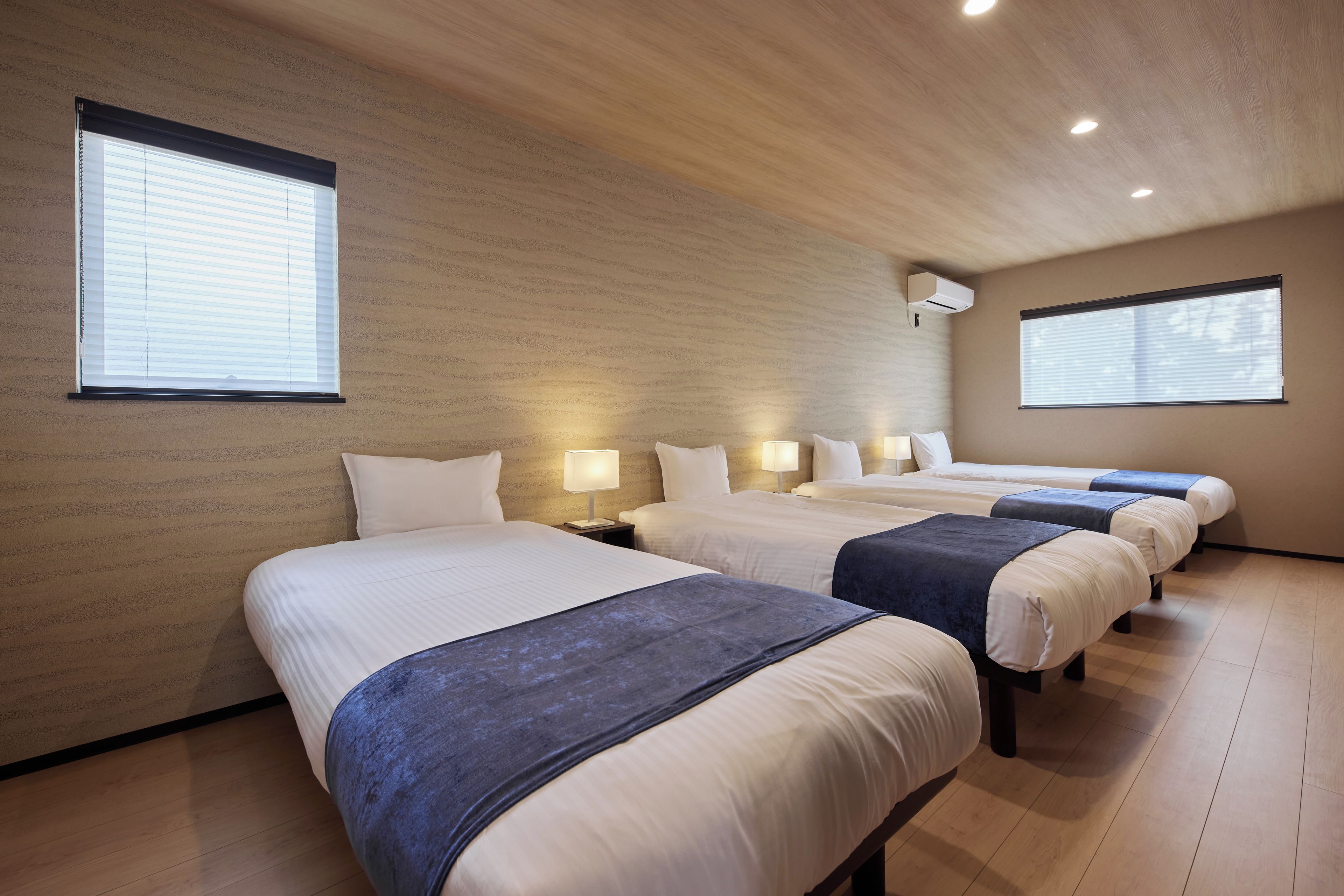 Quarto de hotel com quatro camas, cada uma com cabeceira e mesas de cabeceira, e duas janelas com persianas.