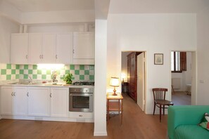 Cozinha compacta com armários brancos, fogão e pia. Na área de estar adjacente, há uma mesa pequena com abajur e uma cadeira.