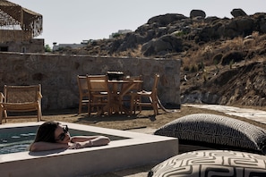 Uma área de piscina com alguém relaxando, móveis de madeira e uma parede de pedra.