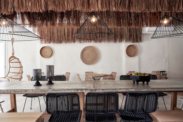 Espaço de refeições com uma mesa comprida, cadeiras de vime preto e decorações de parede em tecido.